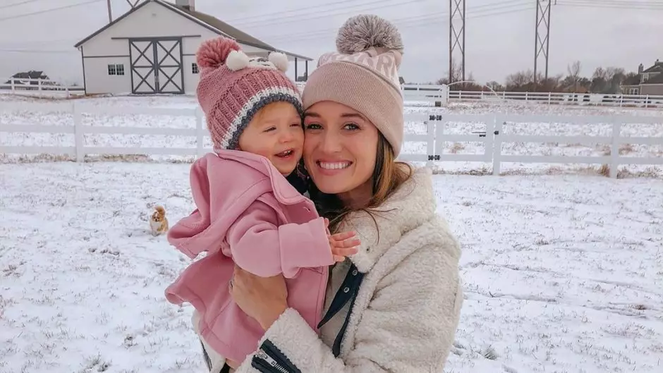 Bachelor Alum Jade Roper Mengenakan Mantel dan Topi Musim Dingin Dengan Puffball di Atasnya Sementara Dia Memegang Putri Emmy dengan Jaket dan Topi Merah Muda