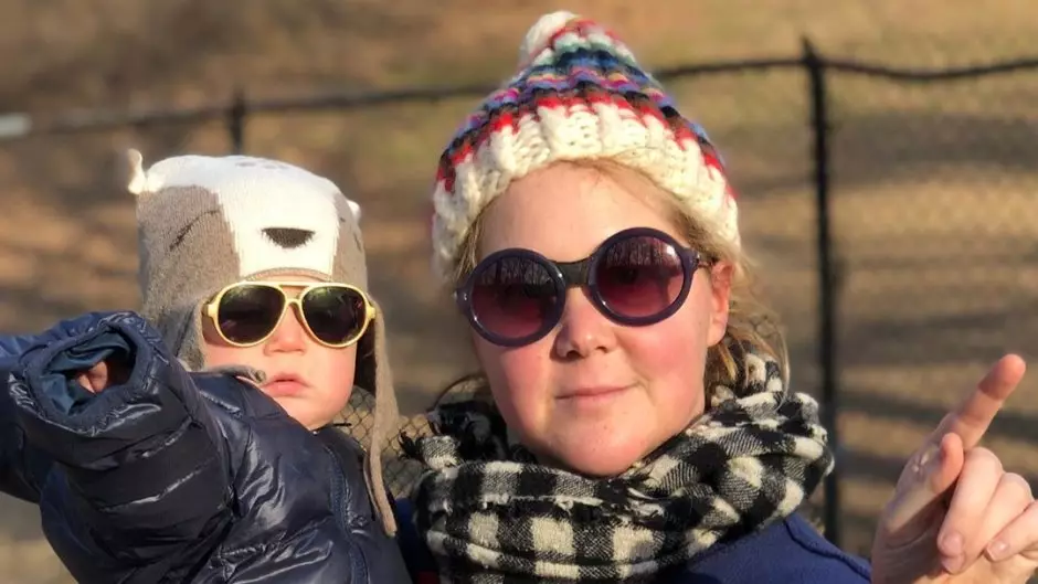Amy Schumer lächelt und trägt eine Sonnenbrille, einen Hut und einen Schal, während sie Sohn Gene in einem Parka-Hut und einer Sonnenbrille hält