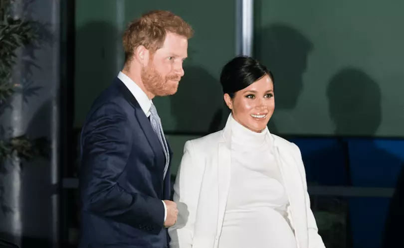 Pangeran Harry nganggo setelan navy karo tangane ing punggung Meghan Markle nalika dheweke nganggo klambi putih lan jaket