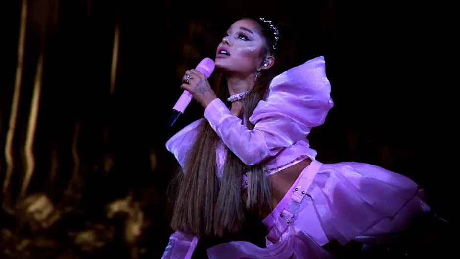 Ariana Grande leži na odru med svetovno turnejo Sweetener v roza kratkem topu in krilu z rožnatim mikrofonom