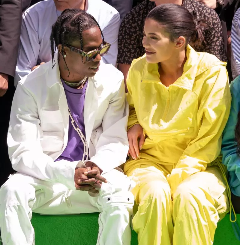 カイリー・ジェンナーは、白いスーツと紫色のシャツを着たトラビス・スコットの隣に座っている間、黄色のジャンプスーツを着ています。