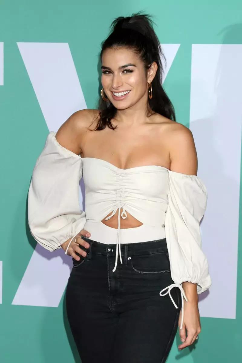 Bachelor Star Ashley Iaconetti brosir í hvítum axlarbol og svörtum gallabuxum