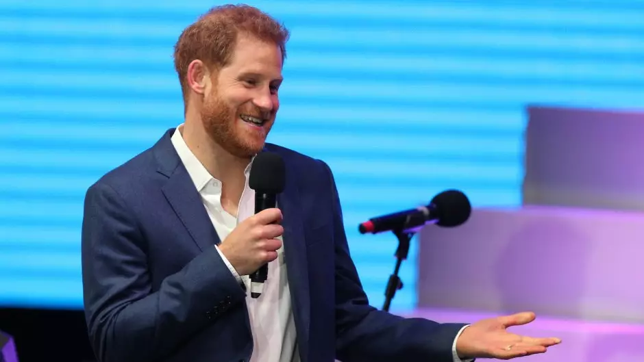 El príncep Harry s'arronsa d'espatlles i somriu amb un blazer blau marí amb camisa blanca amb botons mentre sosté un micròfon