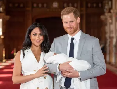 Meghan Markle Souri nan yon rad blan Style trench kanpe bò kote Prince Harry nan yon kostim gri ak kravat ble maren yo ap kenbe ti bebe Archie ki vlope nan rad blan swaddling.